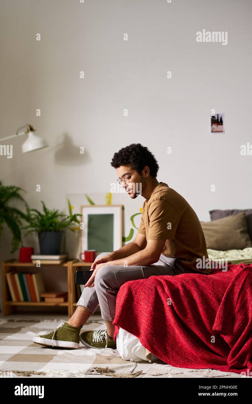 Vertical image of sad teenage boy sitting on bed in his bedroom and ...