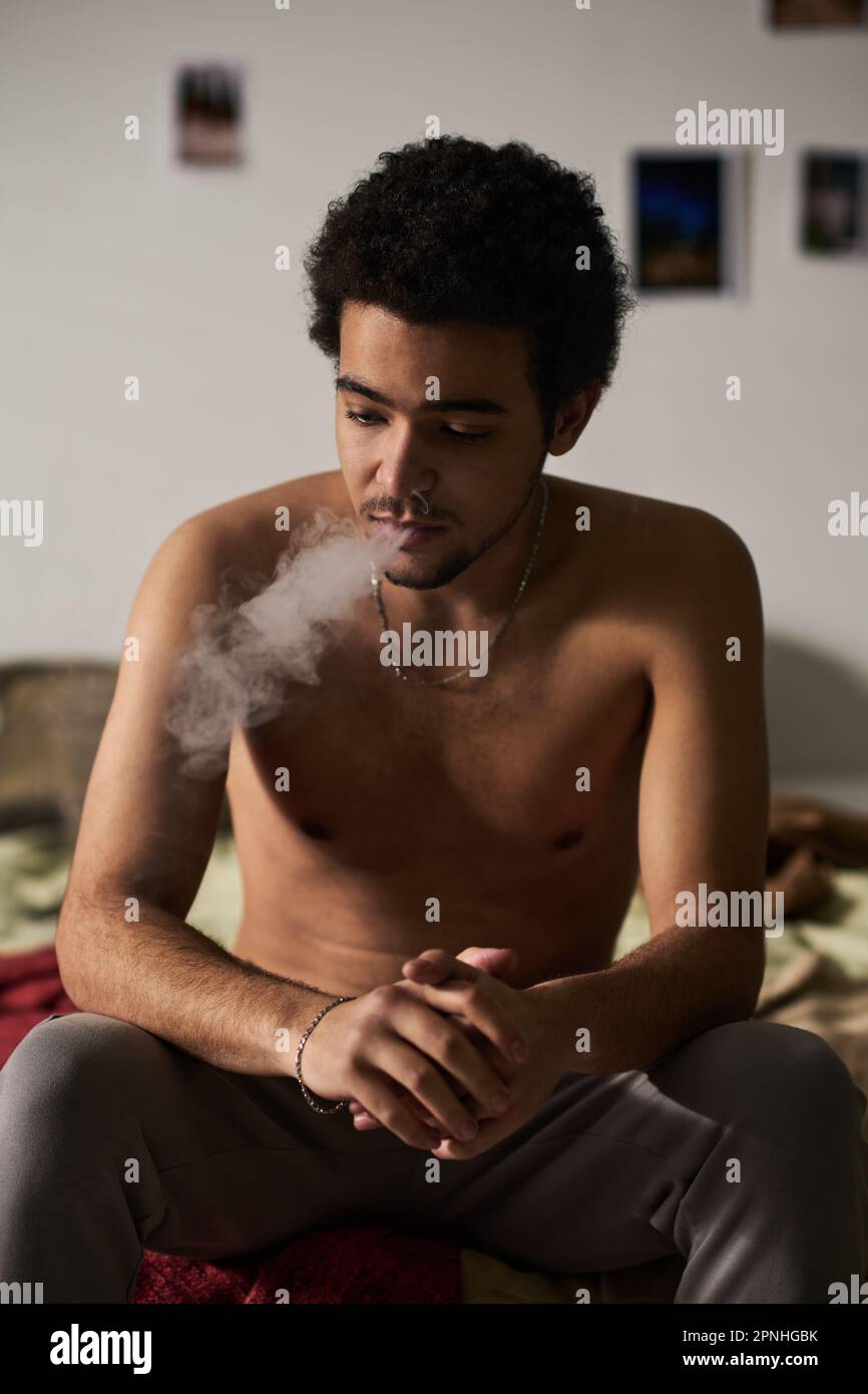 Vertical image of depressed young man smoking while sitting on bed ...