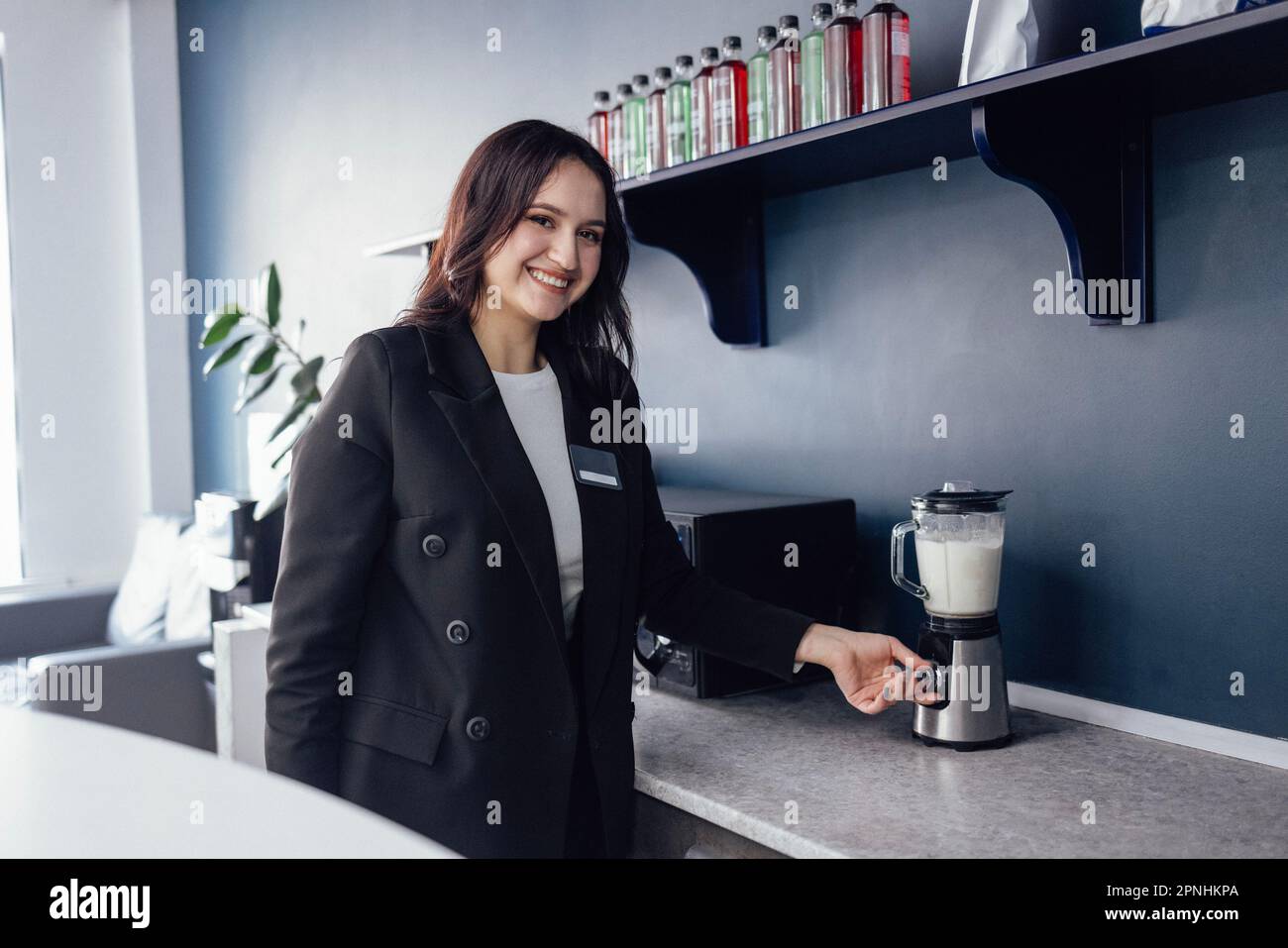 Young pretty woman prepares protein shake using stationary blender. Female barista pours ...