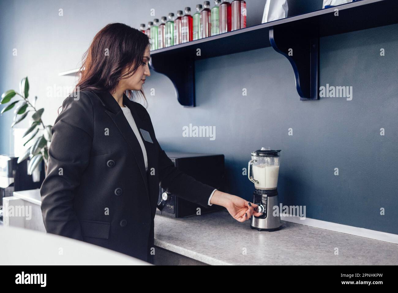 Young pretty woman prepares protein shake using stationary blender. Female barista pours ...