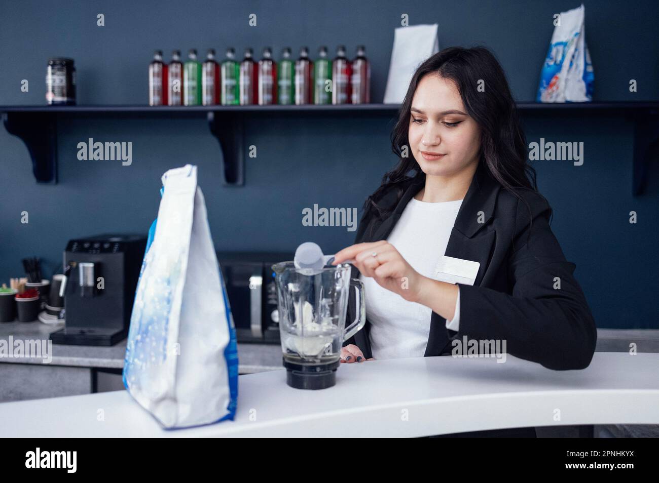 Young pretty woman prepares protein shake using stationary blender. Female barista pours ...