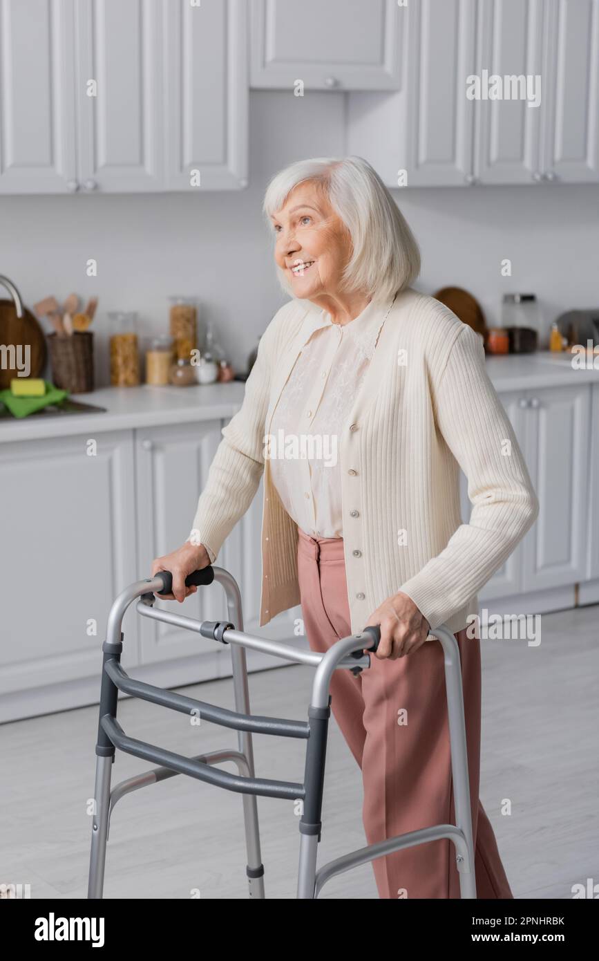 positive senior woman with grey hair walking with help of walker at ...