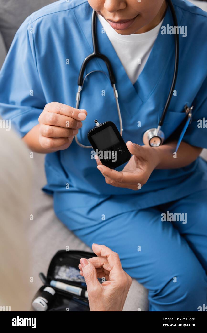 cropped view of multiracial nurse in uniform holding glucometer with ...