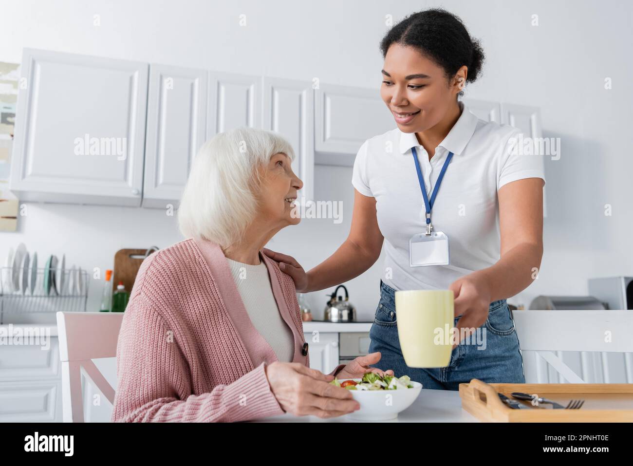 happy multiracial social worker serving lunch to smiling senior woman ...