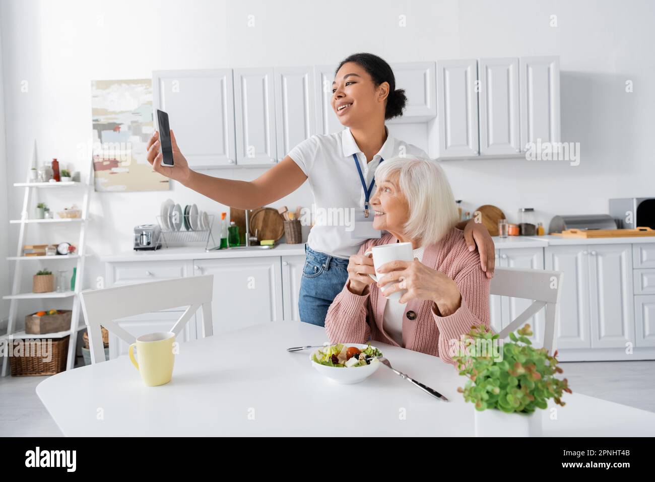 happy multiracial social worker taking selfie with retired woman during ...