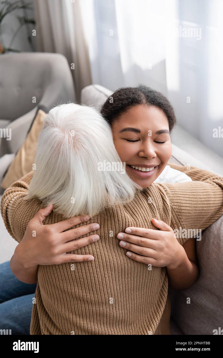 happy multiracial social worker with closed eyes hugging with senior ...