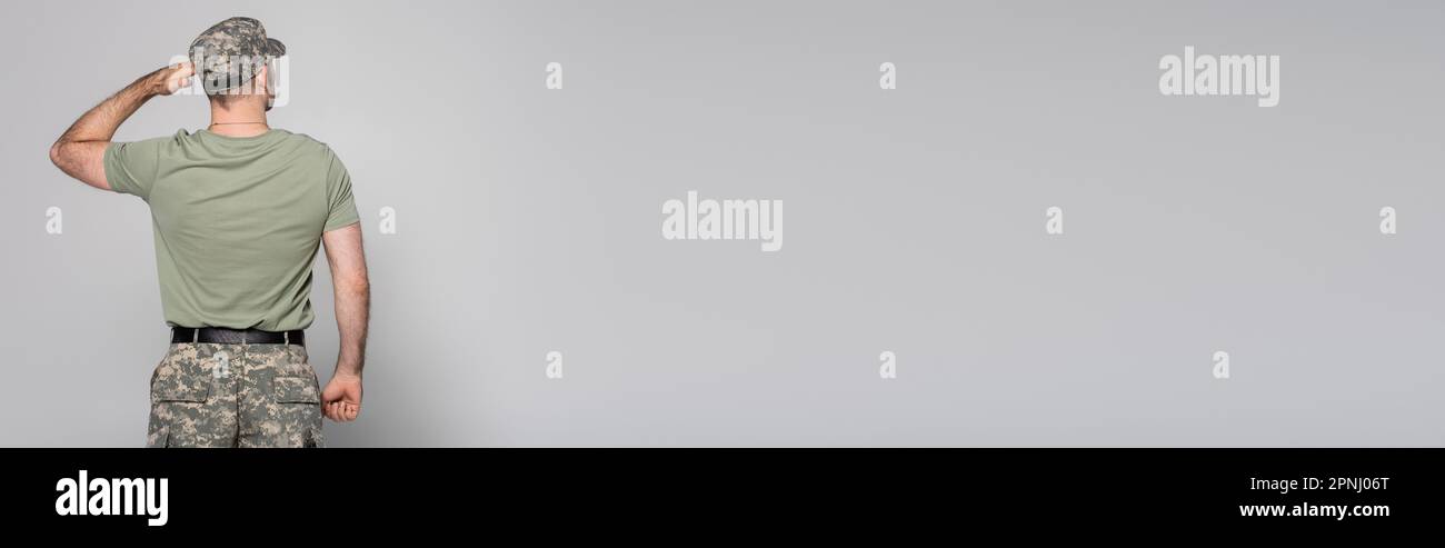 back view of serviceman in military uniform and cap saluting on grey ...