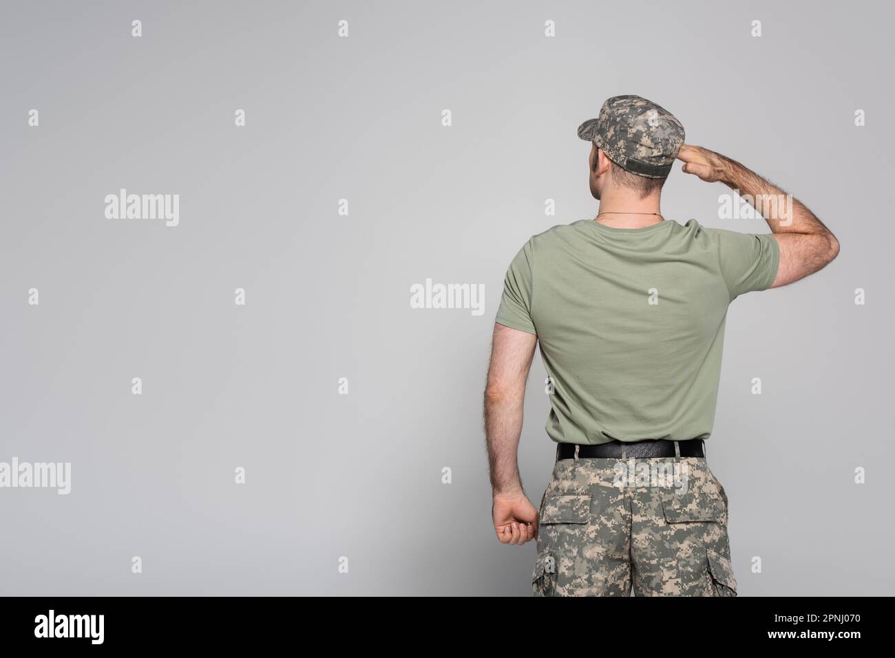 back view of serviceman in military uniform and cap saluting on grey ...