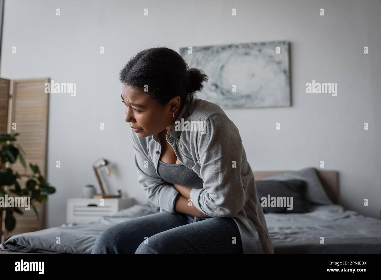 Tired multiracial woman feeling cramp in stomach while sitting on bed ...