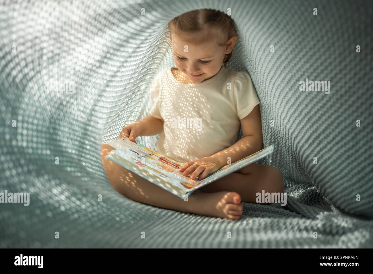 Little girl enjoying of review a book under blue knitted plaid in sunny ...
