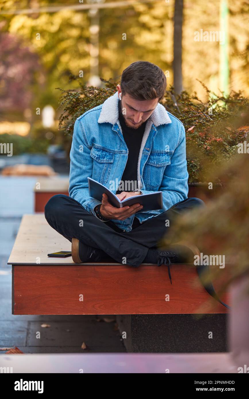 A man sitting on a bench in the city center reading a book while ...