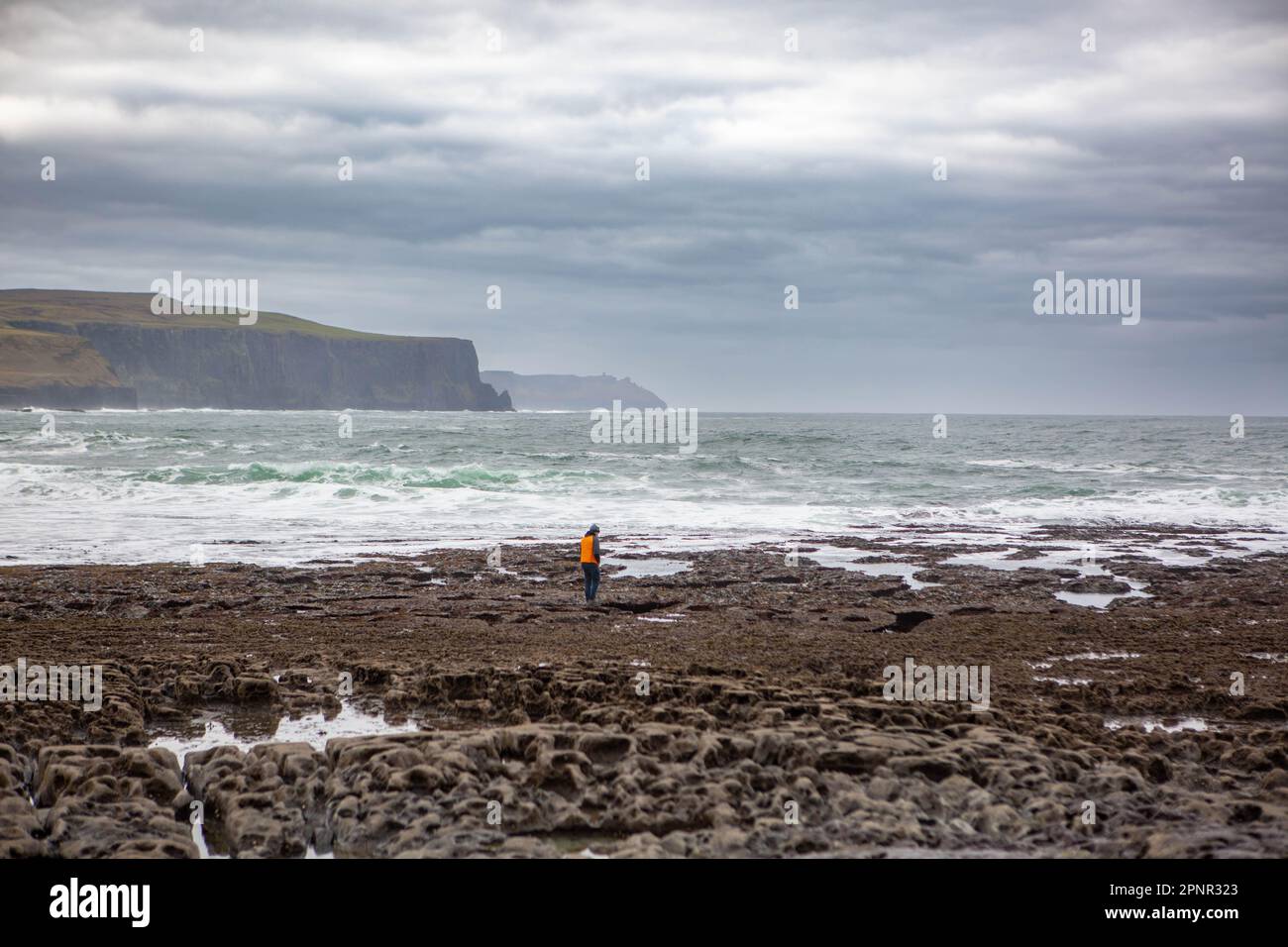 Distant rear view of a man walking on a rocky beach by Cliffs of Moher ...