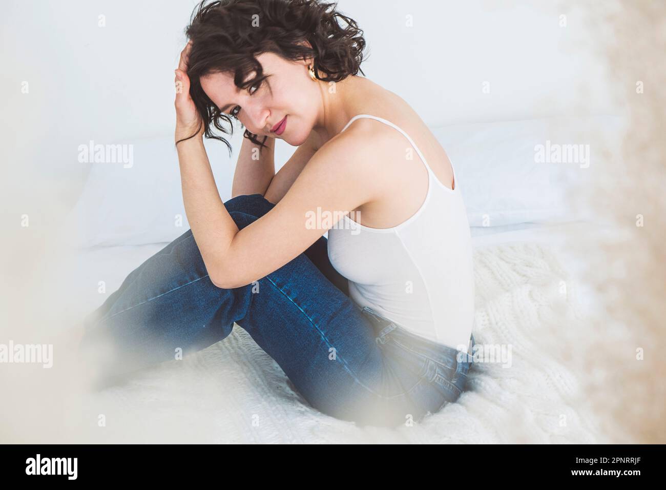 A brunette with curly hair is sitting on the bed and hugging her knees ...