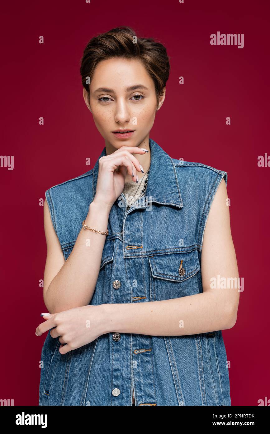 young model with short hair posing in denim vest on dark red background ...