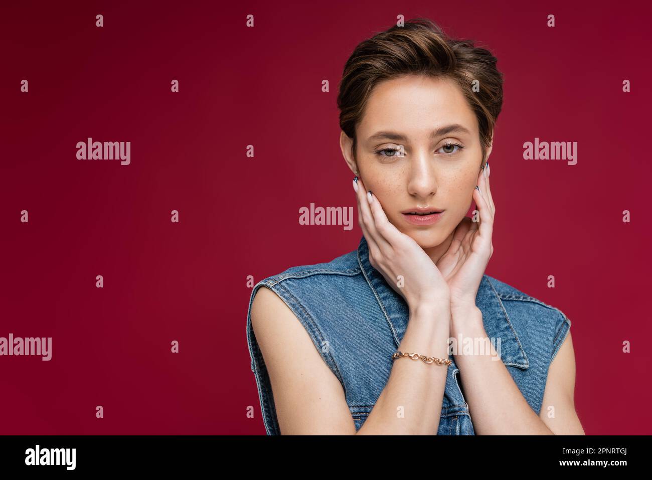 stylish model in denim outfit with vest touching face on burgundy ...