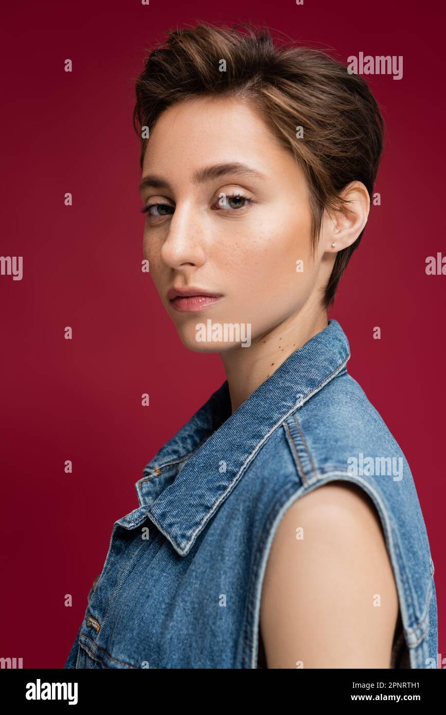 charming young model with short hair posing in denim vest on burgundy ...