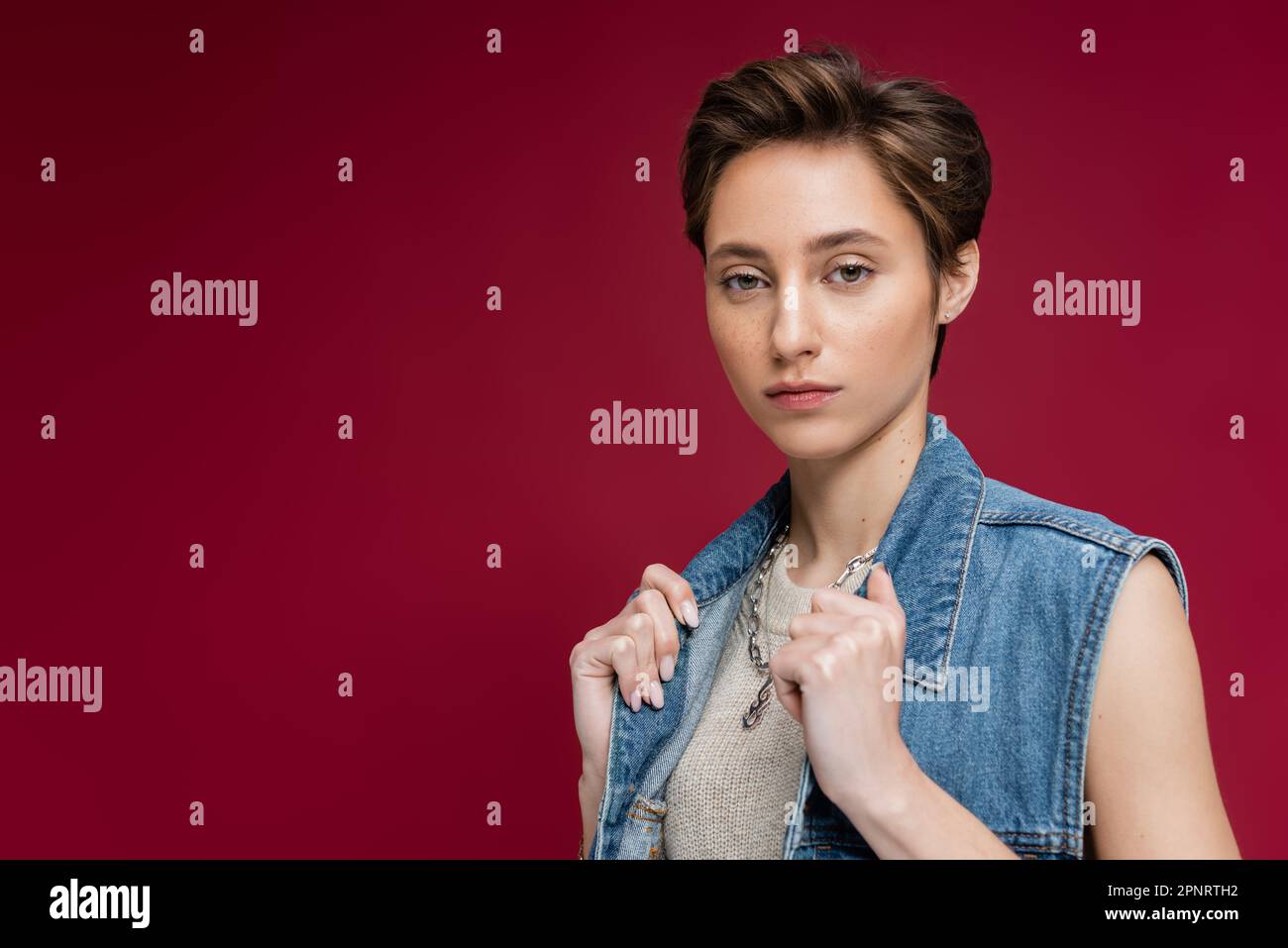 stylish model in adjusting denim vest while posing on burgundy ...