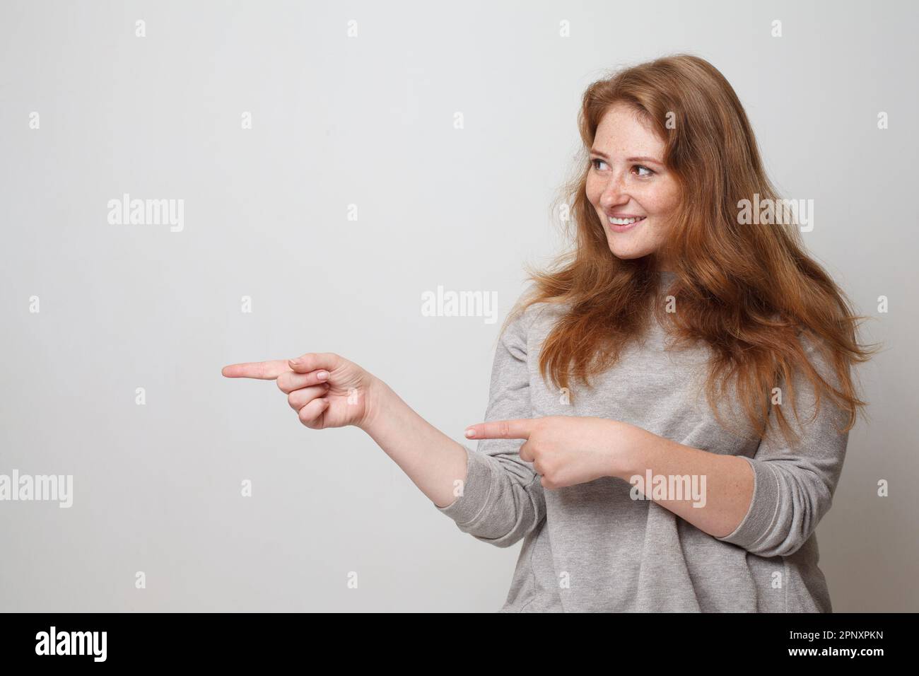 Young smiling woman pointing finger aside on white studio wall ...