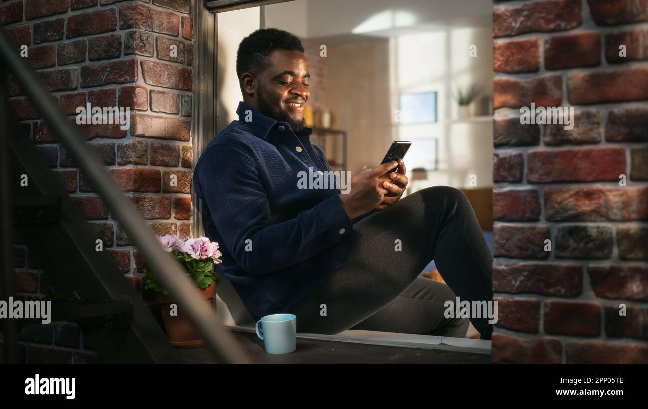 Black Authentic Man Using Smartphone in his Flat While Sitting on Bedroom Windowsill. African ...