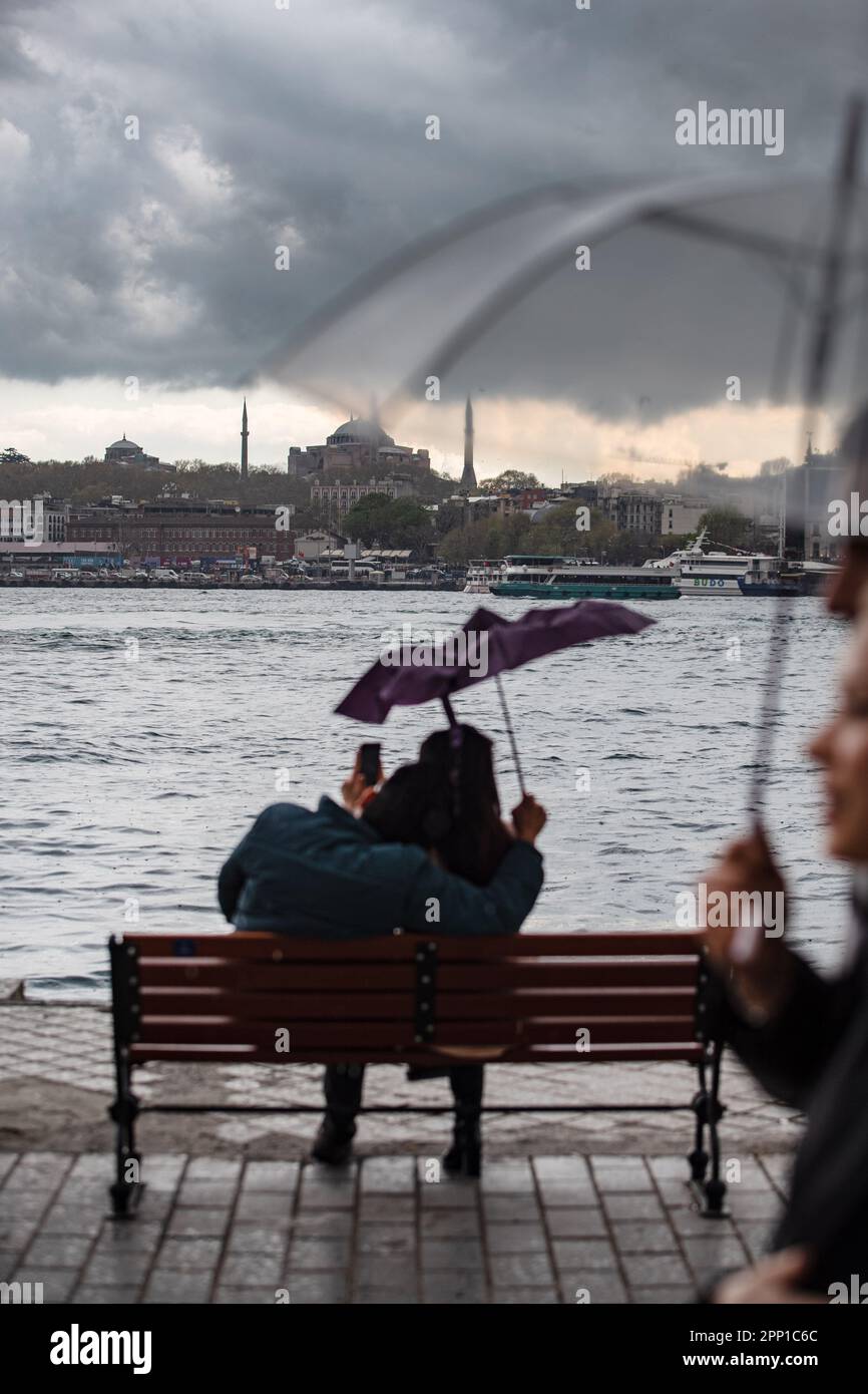 People sitting on the bench on the Karakoy beach hold an umbrella to avoid getting wet from the ...