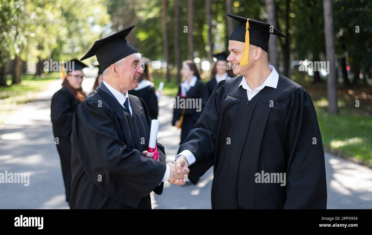 A group of graduates in robes outdoors. An elderly man and a young guy ...