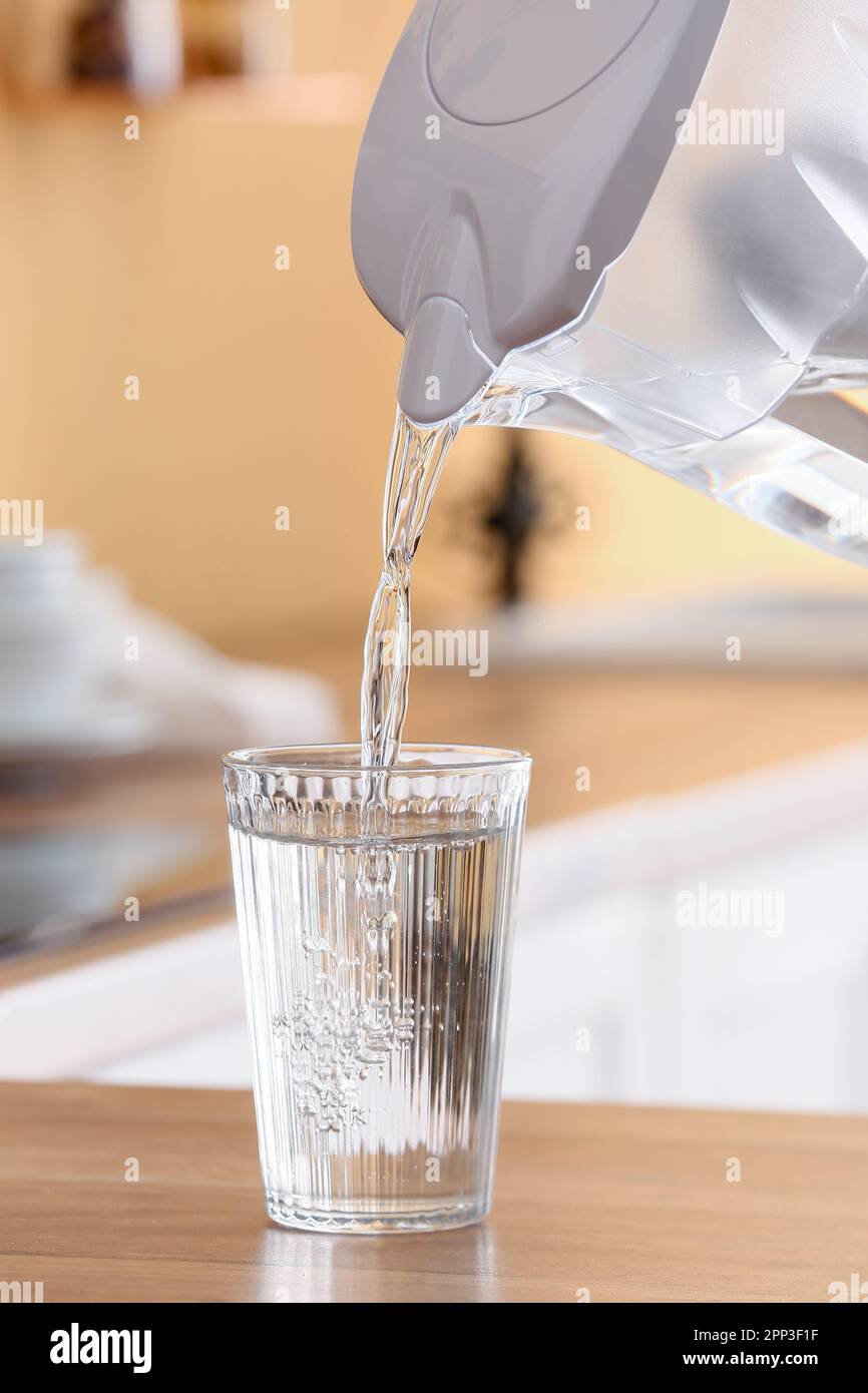 Pouring of water from modern filter jug into glass on kitchen counter ...