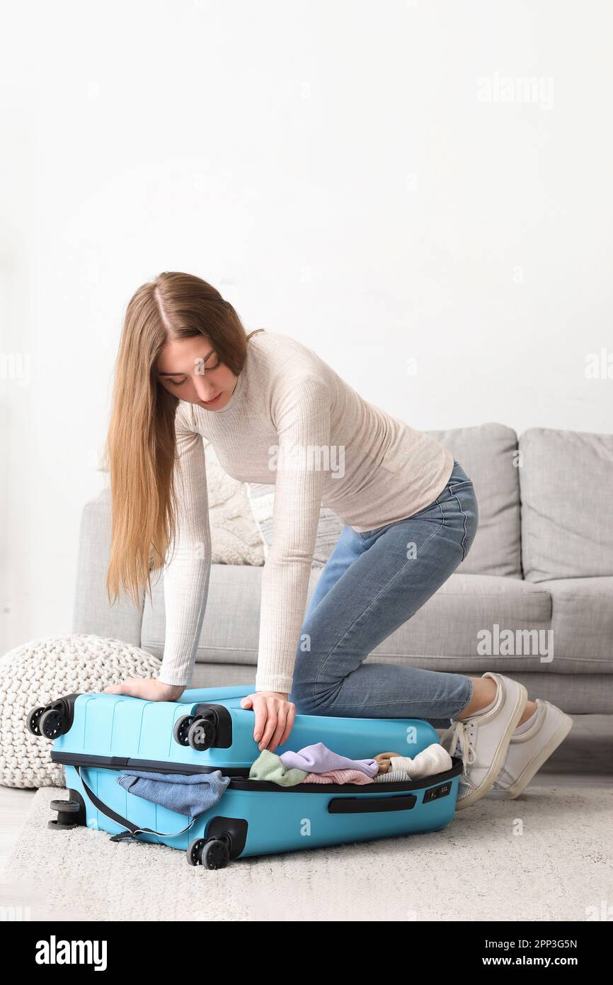 Young woman trying to close suitcase with a lot of clothes at home ...
