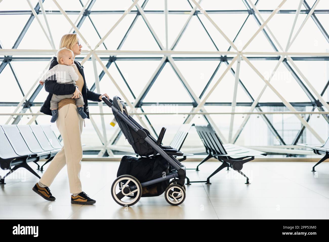 Mother carying his infant baby boy child, pushing stroller at airport ...
