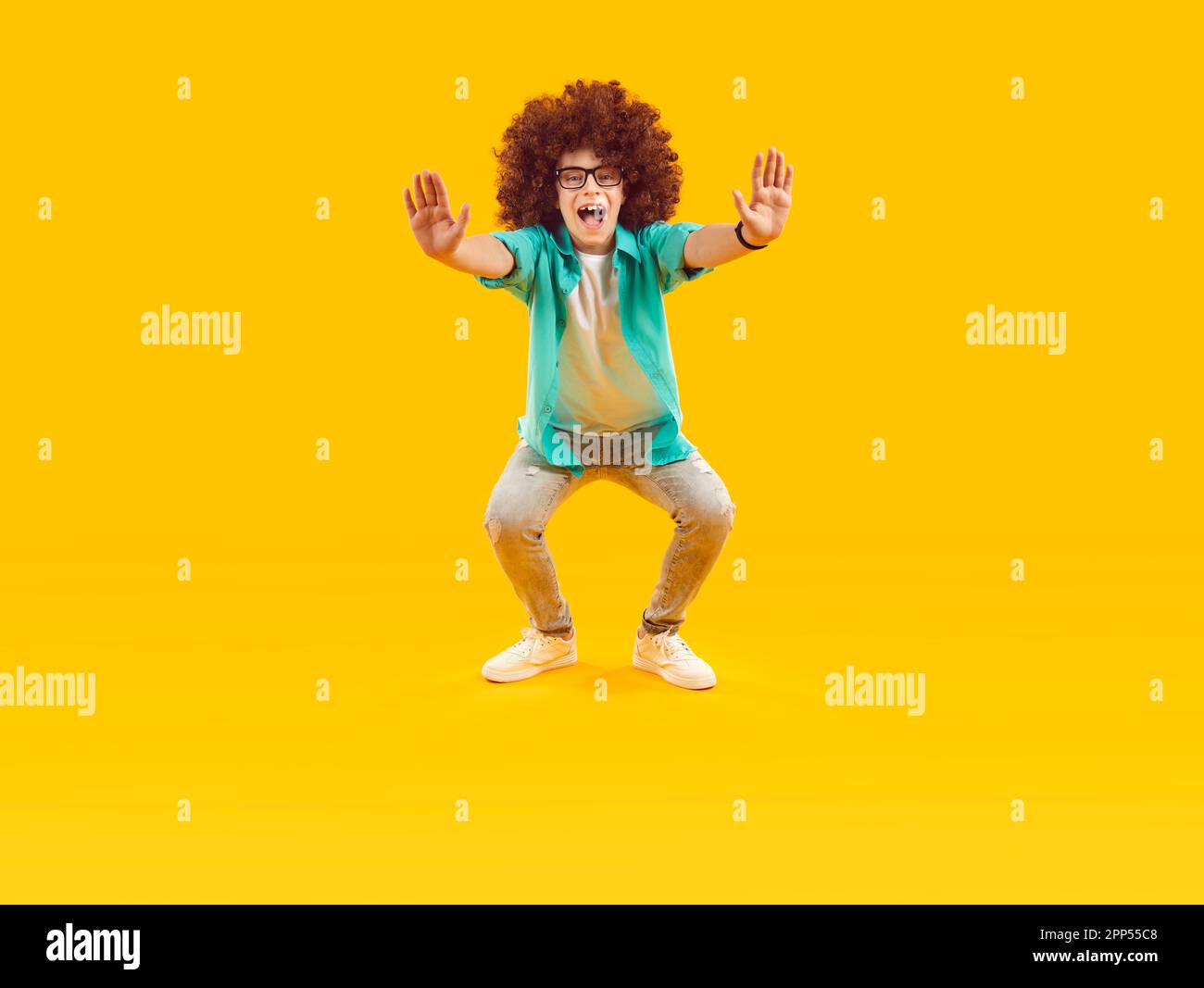 Happy funny little child in curly wig doing squats on orange color ...