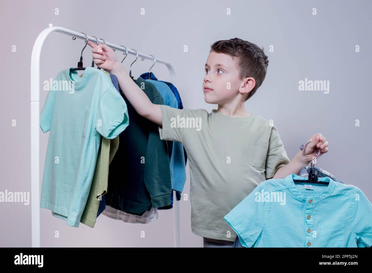 A stylish little preschool boy is standing by the clothes rack, looking ...