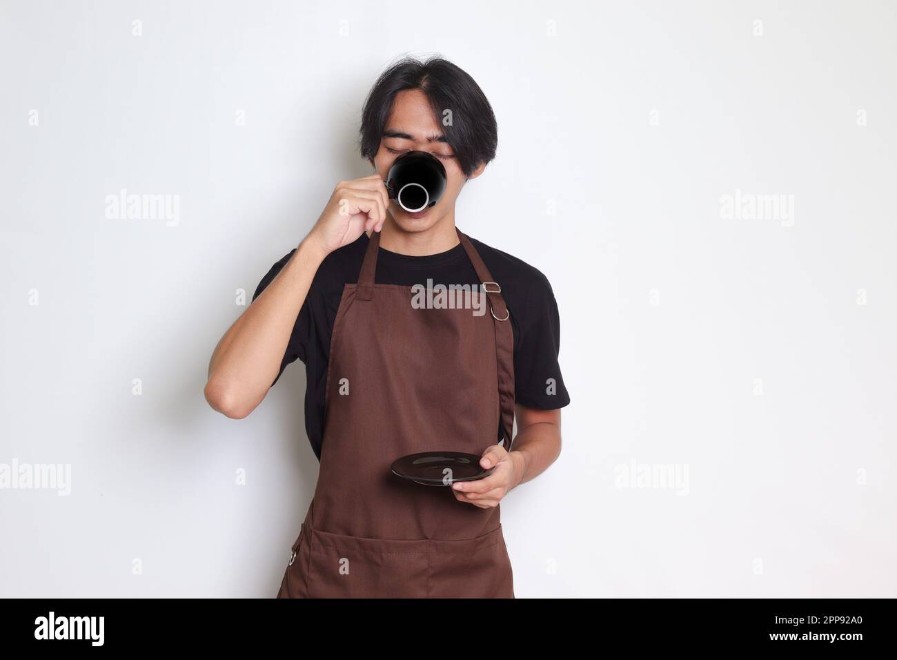 Portrait of attractive Asian barista man in brown apron drinking a cup ...