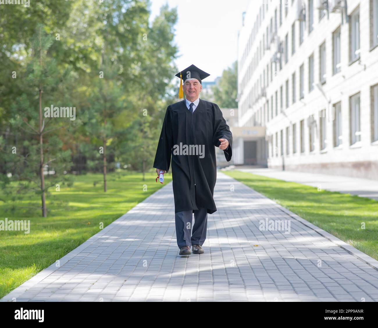 An old man in a graduation gown walks outdoors and holds a diploma ...