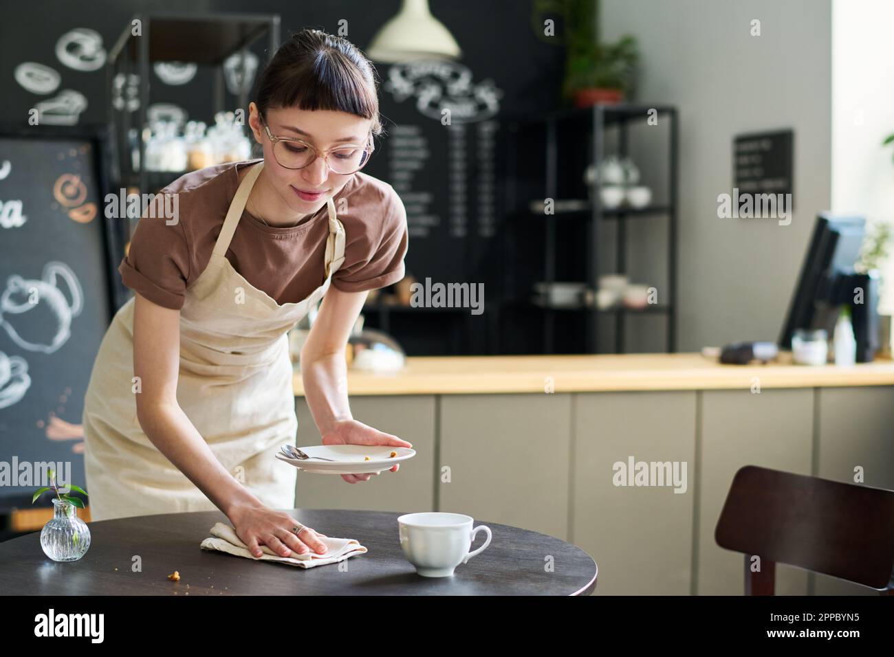 Young pretty waitress in apron and eyeglasses holding plate with spoon ...