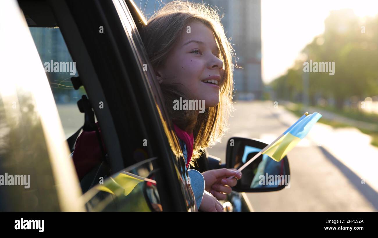 Caucasian girl in car window with Ukrainian flag in hand. Kid smiles at ...