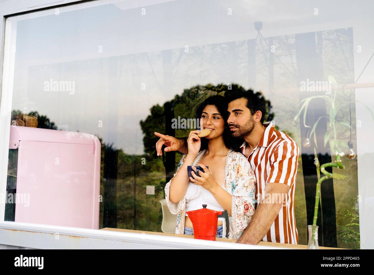 Man pointing by woman eating biscuit seen through window glass at home ...
