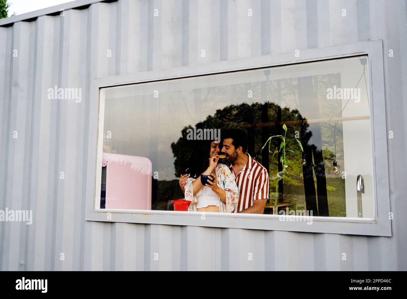 Woman feeding boyfriend in kitchen seen through window of container ...