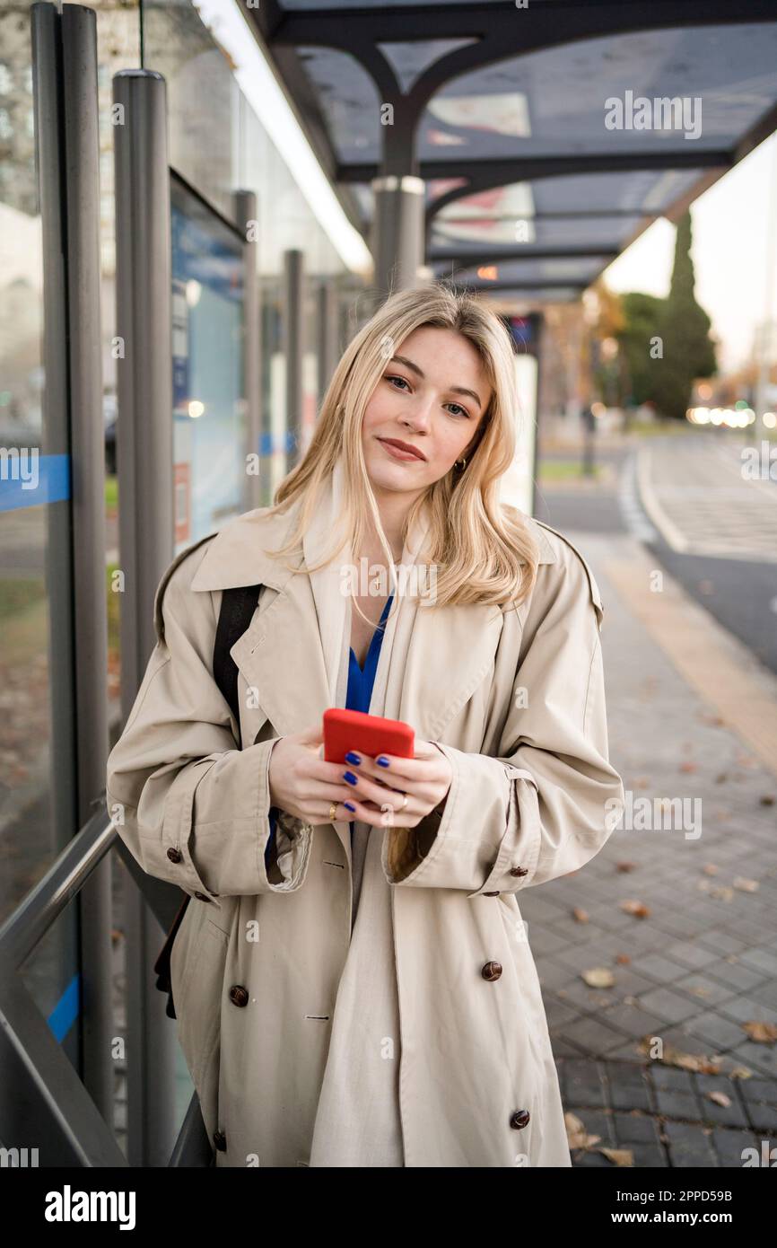 Young woman holding smart phone standing at bus stop Stock Photo - Alamy