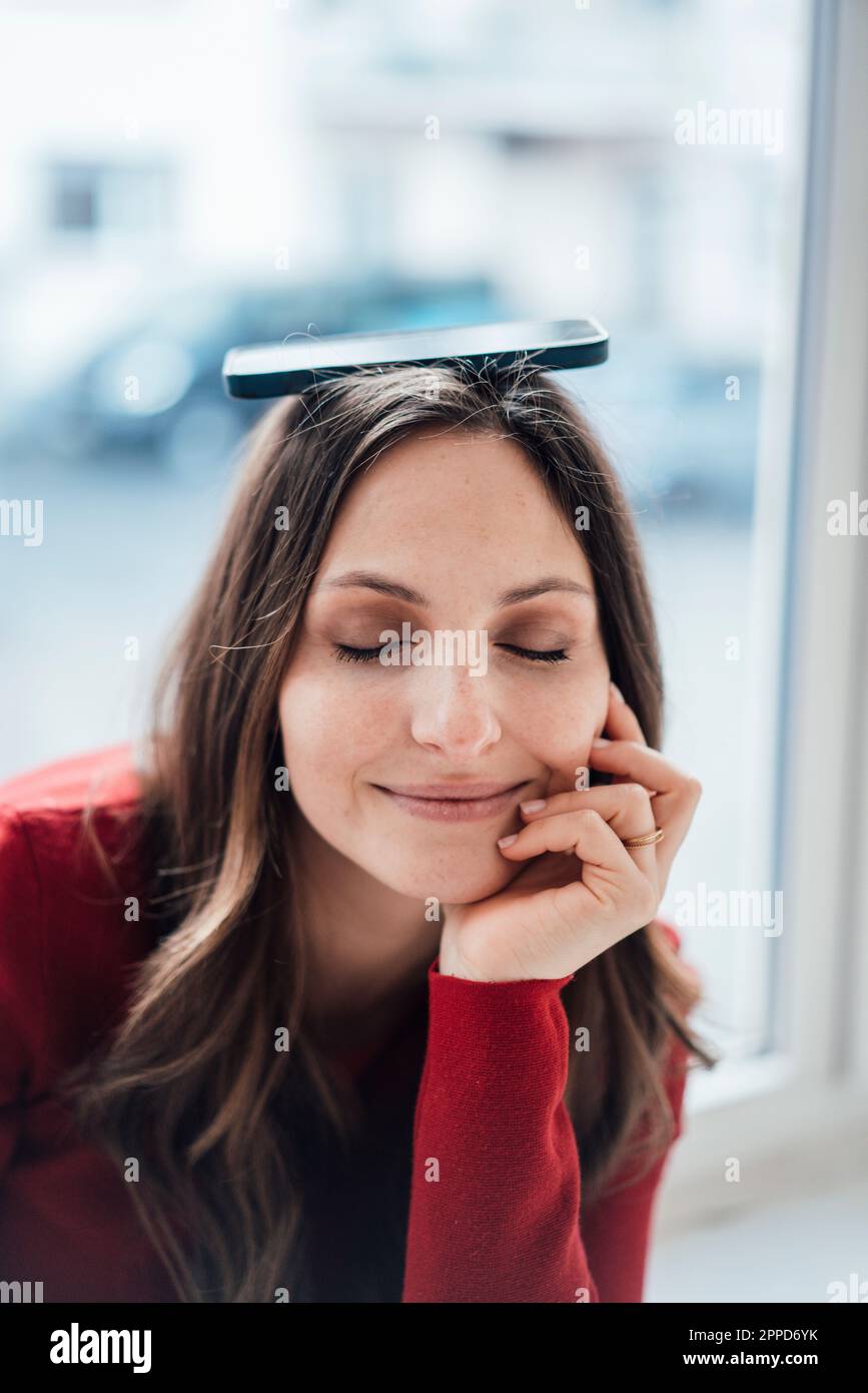 Woman with eyes closed balancing smart phone on head in front of window ...