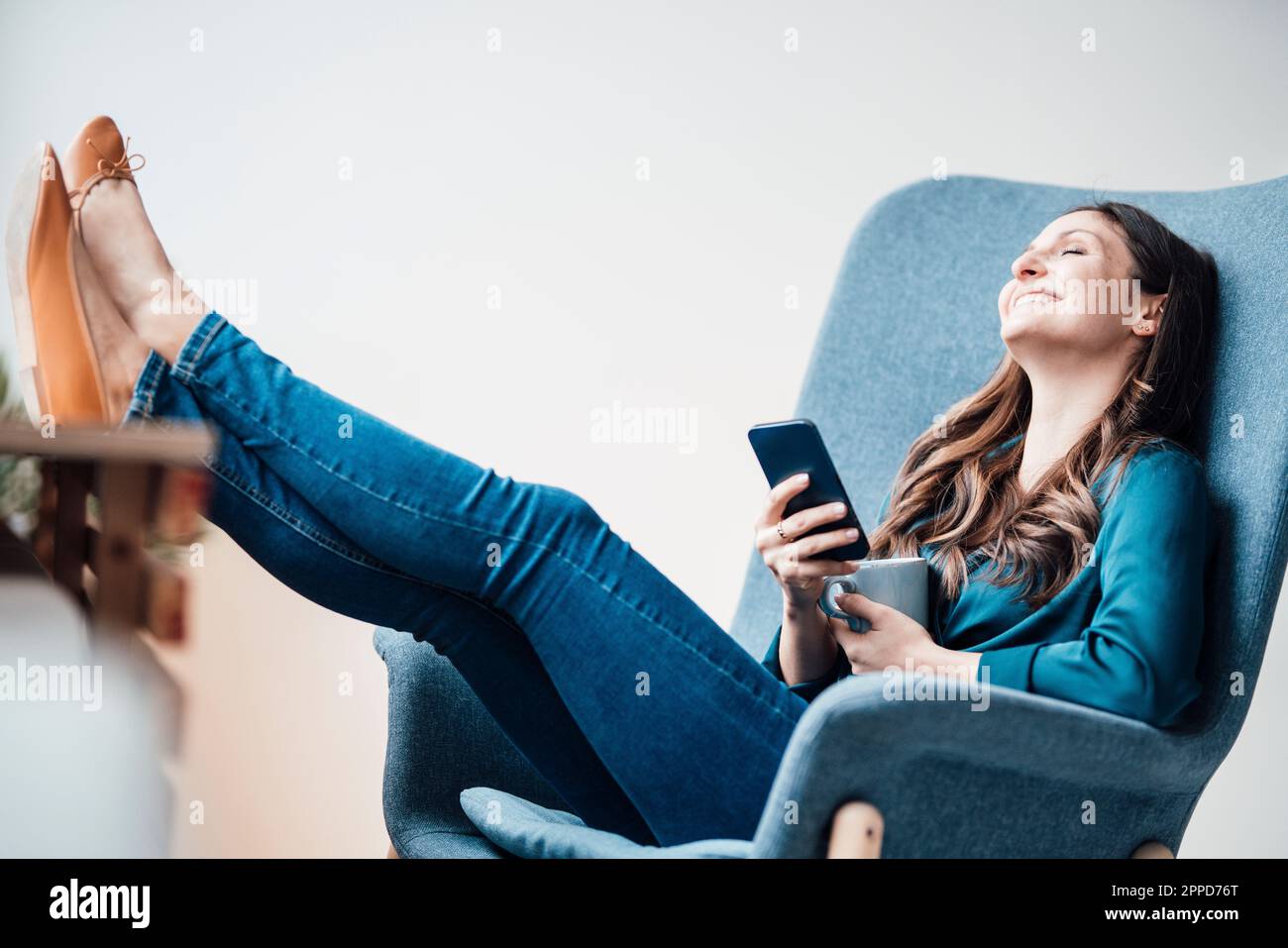 Happy young woman with smart phone relaxing in armchair with feet up at ...