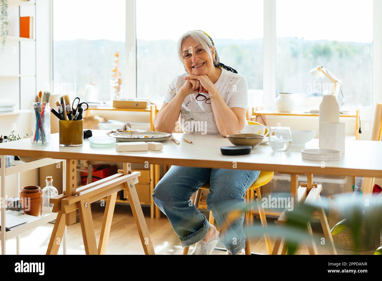 Happy senior woman sitting with hand on chin at table in workshop Stock ...