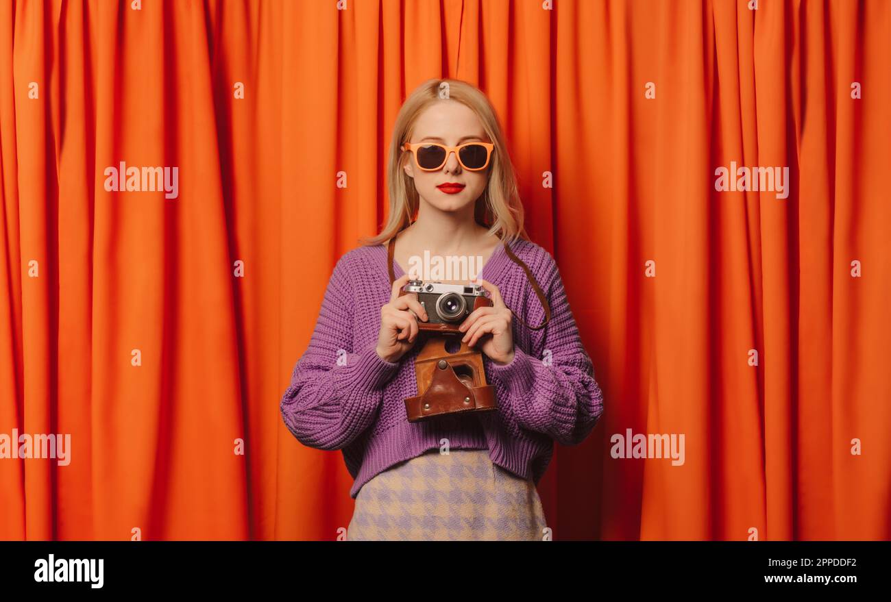 Blond woman with vintage camera standing in front of orange curtain ...