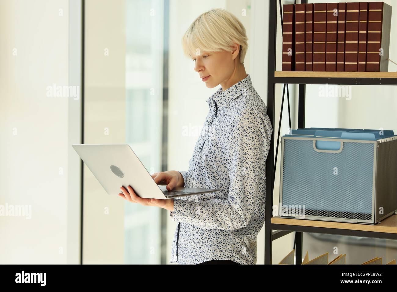 Female employee works on laptop leaning on rack in office Stock Photo ...