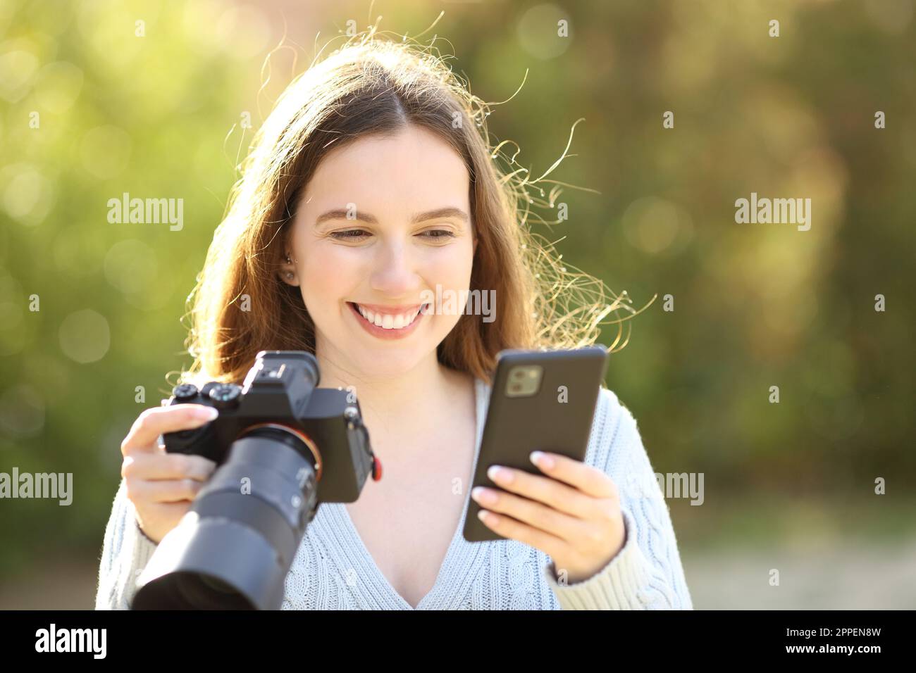 Happy photographer checking phone holding mirrorless camera in a park ...