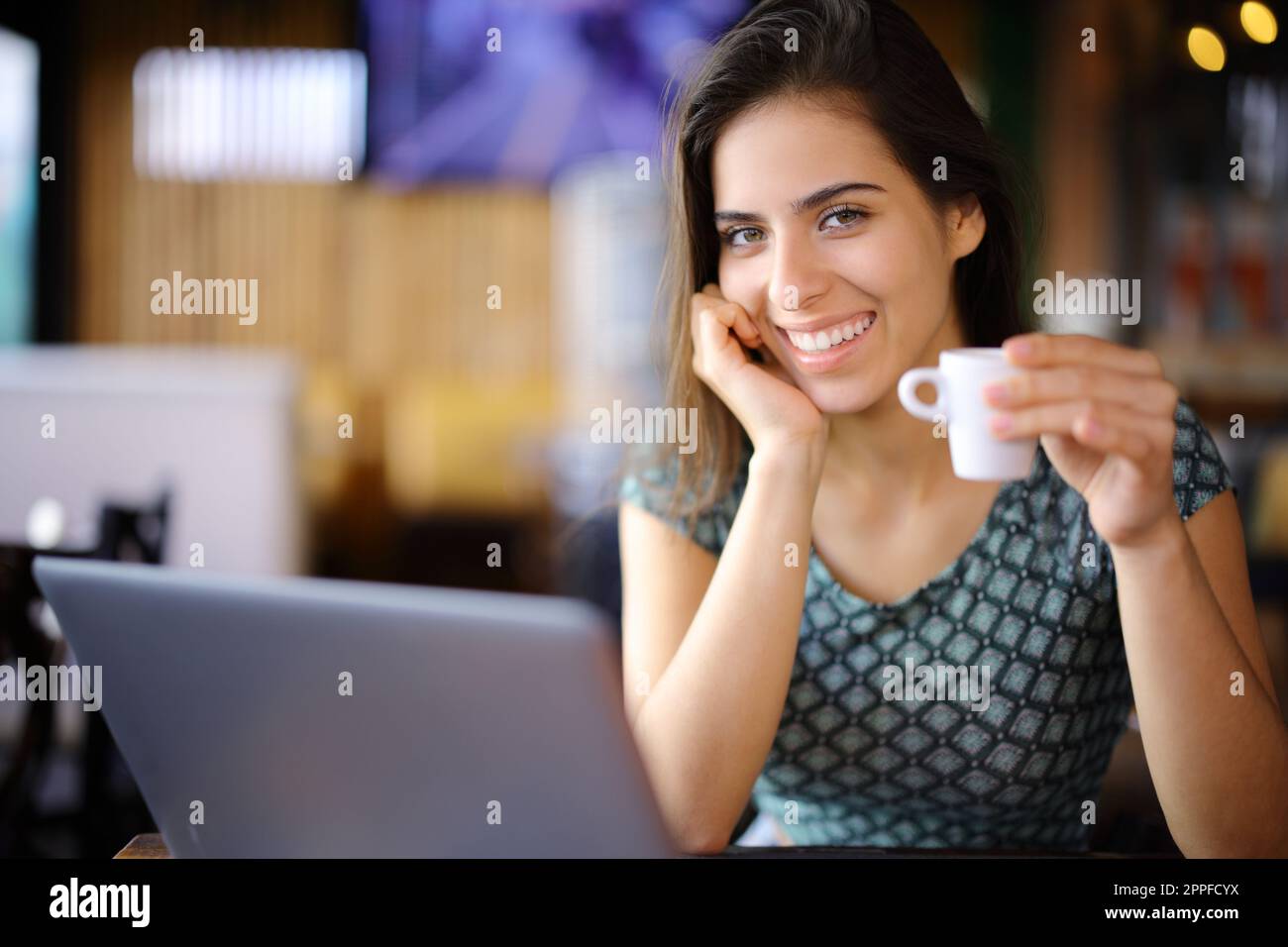 Happy woman with laptop drinking in a coffee shop looking at camera ...