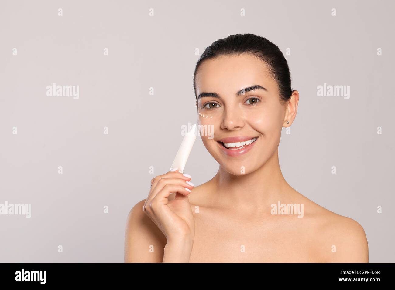 Beautiful young woman applying gel on skin under eye against light grey ...