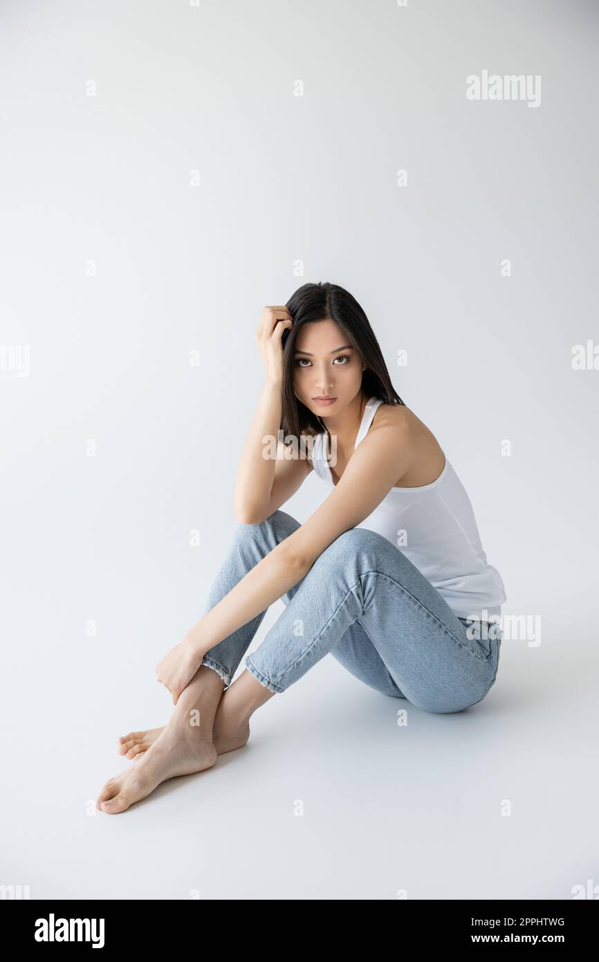 Full Length Of Barefoot Asian Woman In White Tank Top And Jeans Sitting And Looking At Camera On