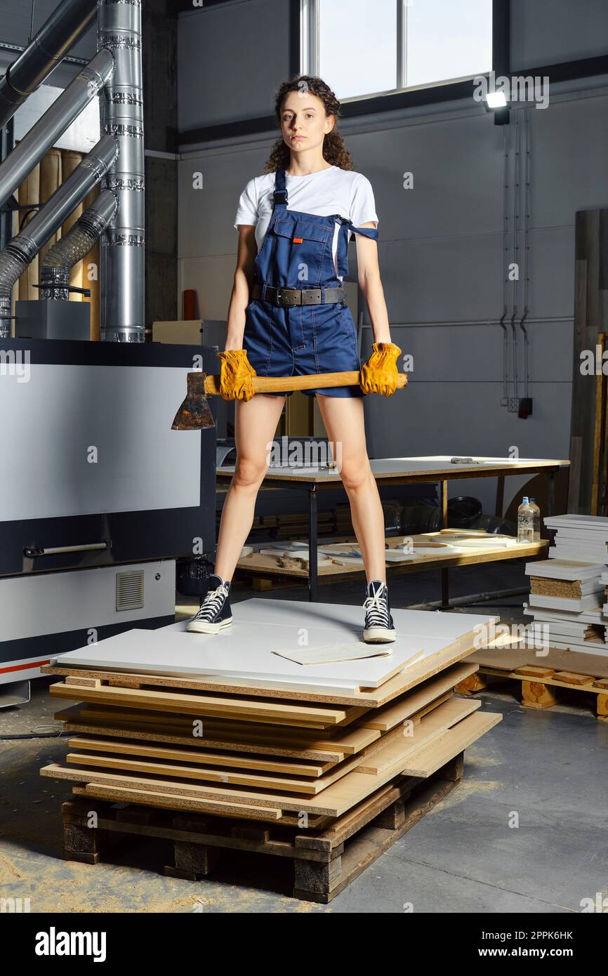 Woman in work clothes stands on a stack of chipboard slabs with an axe ...