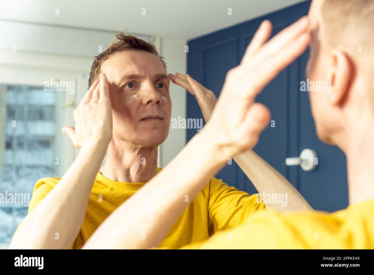 Middle aged man apply cream on clear face, then massage with hands. Male portrait in room mirror ...