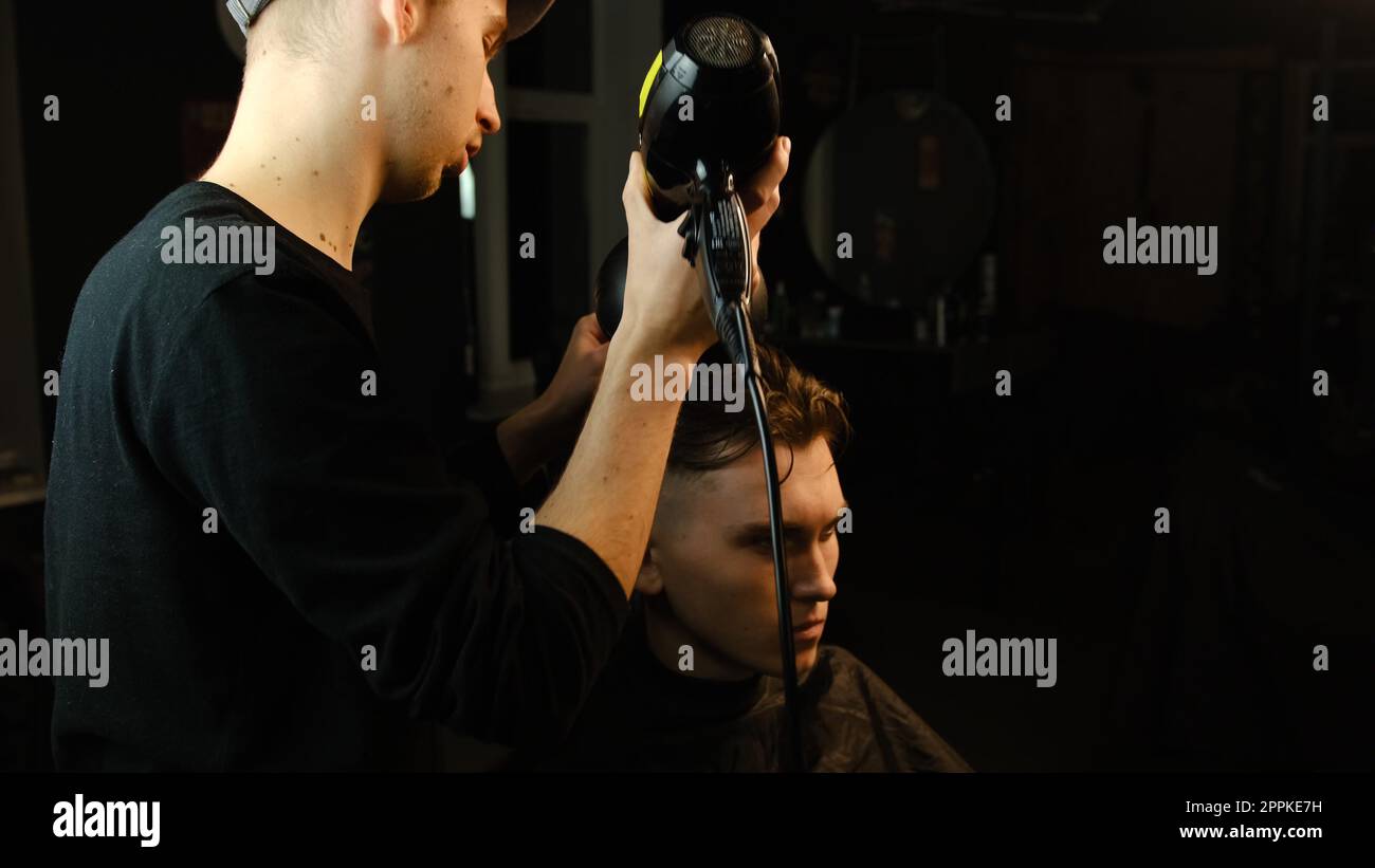 Young barber drying hair of customer with hair dryer in barber shop in ...