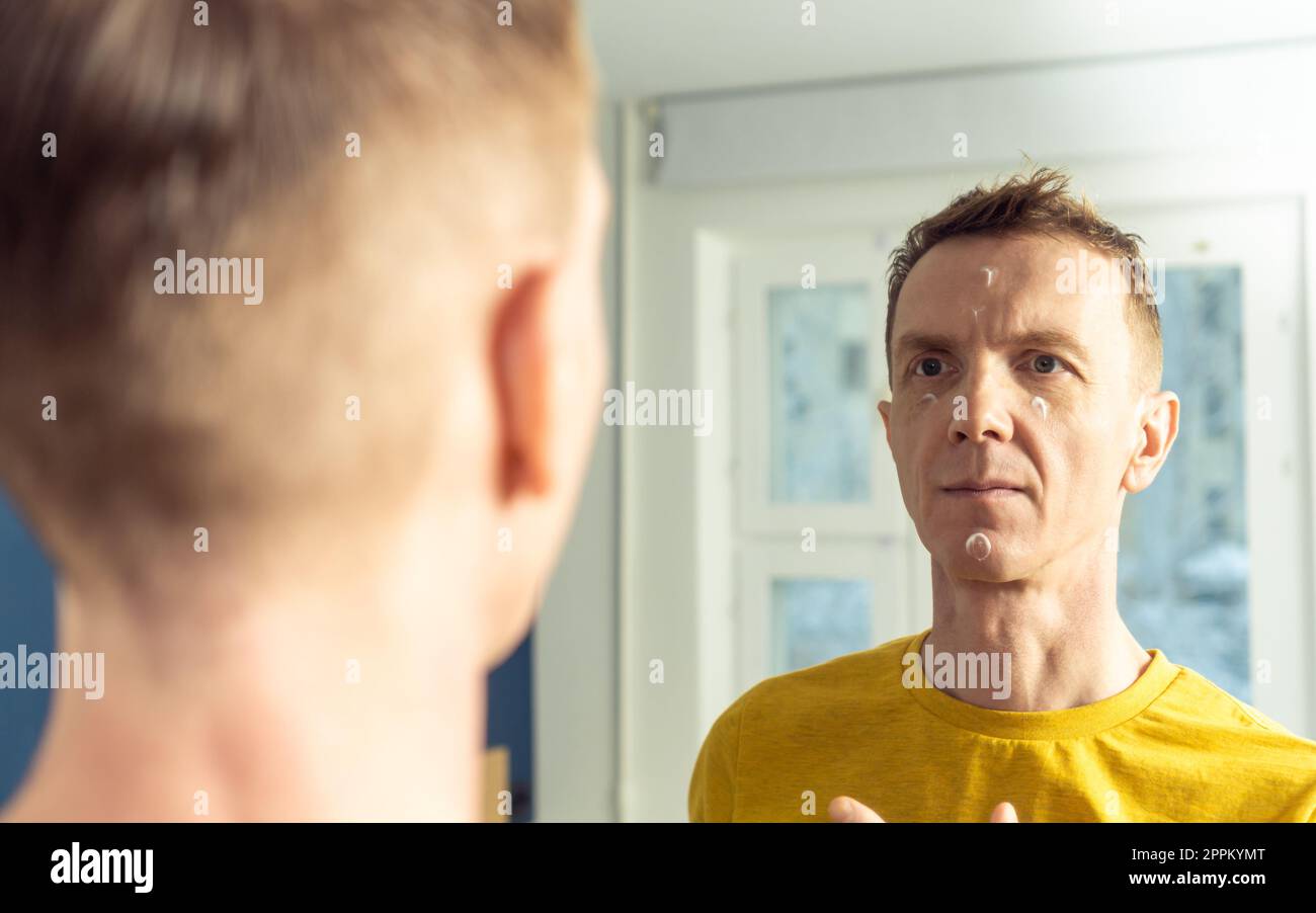 Middle aged man apply moisturizing cream on clear facial areas. Male portrait in room mirror ...